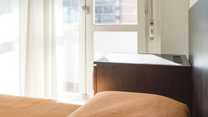 Quarto de hotel com uma cama, uma cômoda de madeira e uma janela com vista para edifícios.