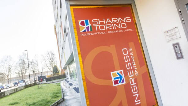 Entrada de um prédio com uma placa indicando 'SHARING TORINO', que oferece moradia social, residência e serviços de hotel.