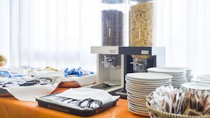 Disposição de buffet com dois grandes recipientes de comida, uma máquina de café e pilhas de pratos organizadas.