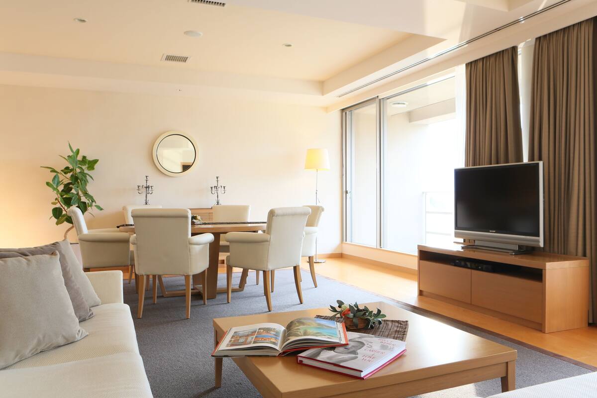 Quarto de hotel moderno com sofá, mesa de centro, televisão, área de jantar e janelas amplas.
