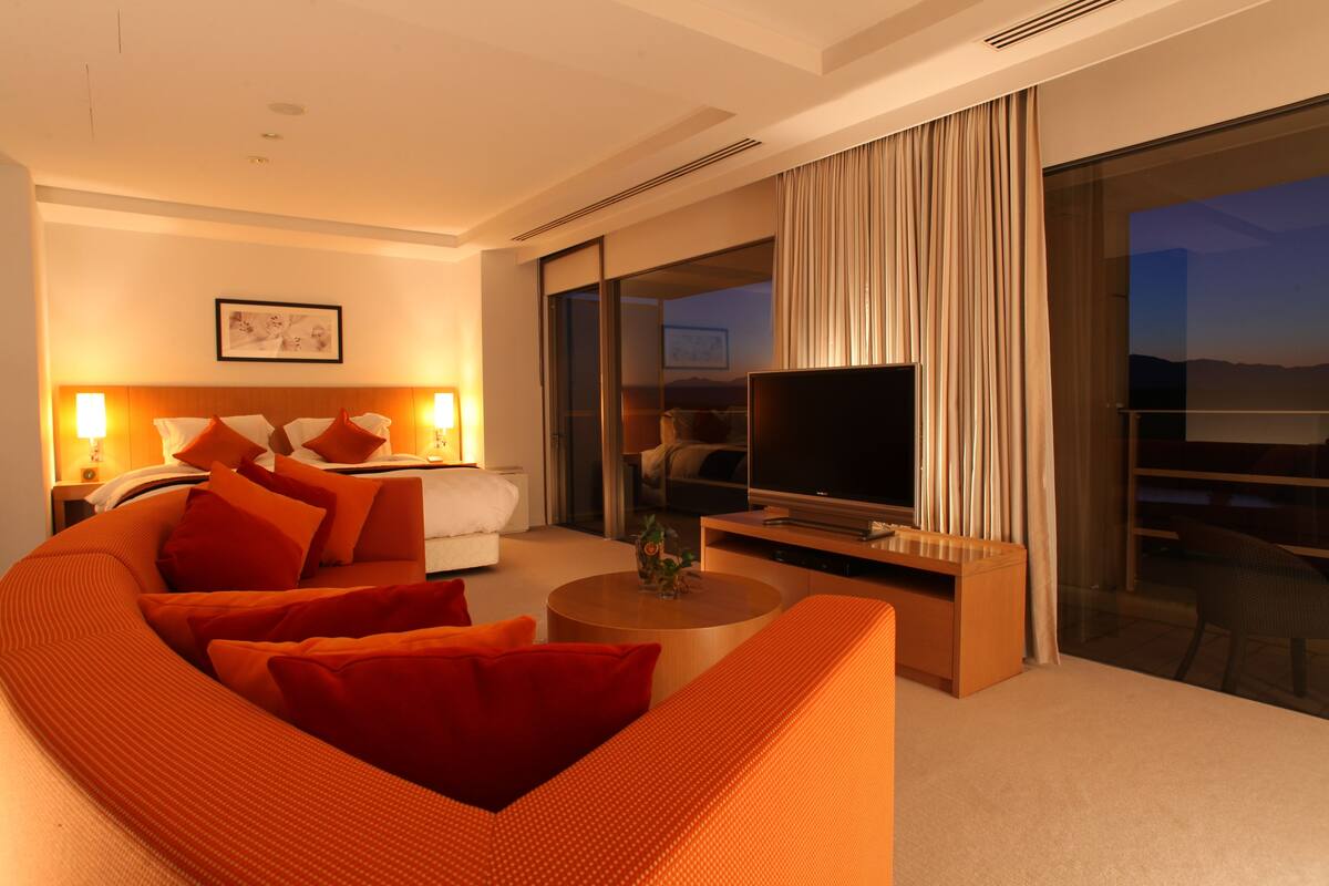 Quarto de hotel moderno com sofá laranja curvo, televisão de tela plana, mesa de centro pequena e uma grande porta de vidro deslizante que dá para uma varanda.