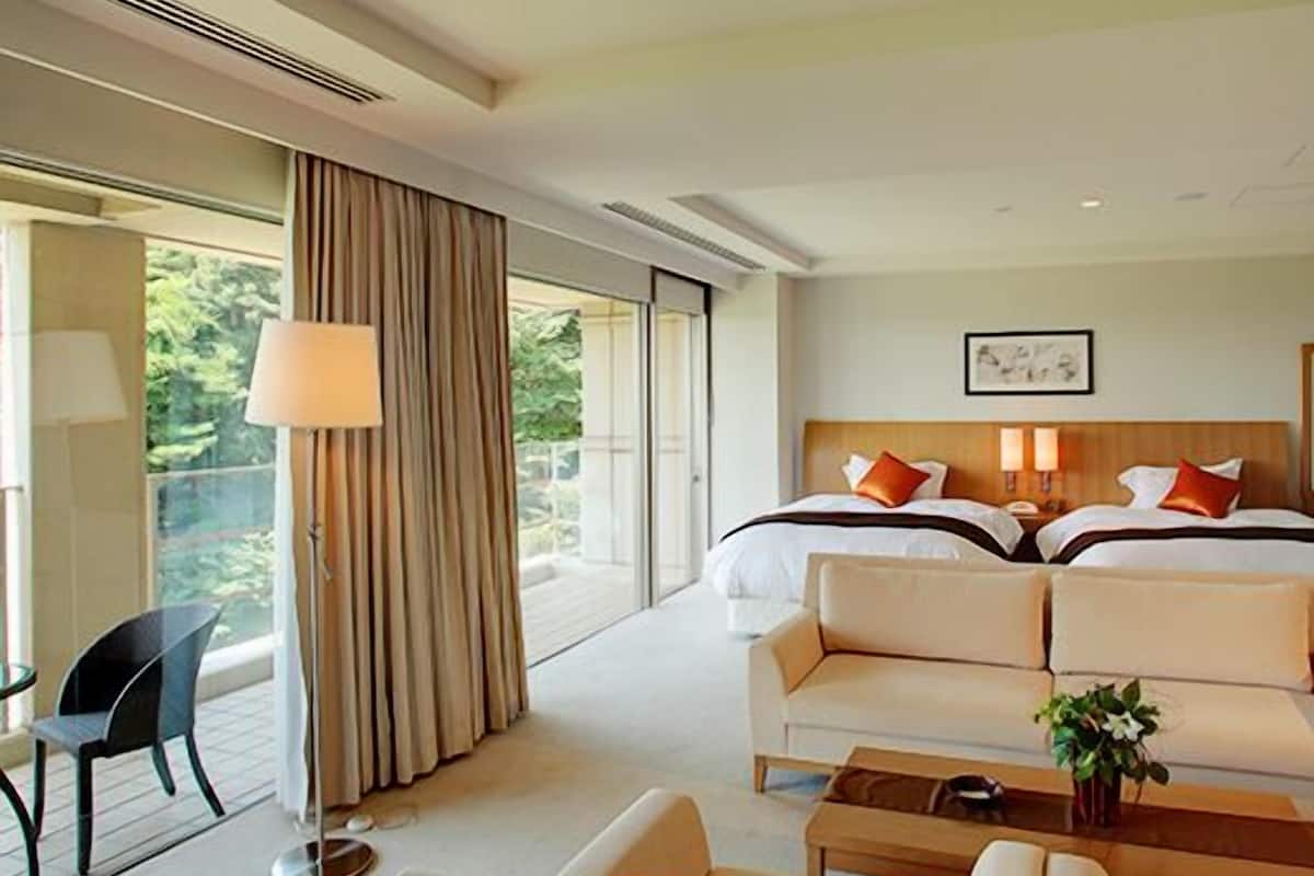 Quarto de hotel moderno com uma cama grande, dois sofás, uma mesa de centro e vista para o verde através de grandes janelas.