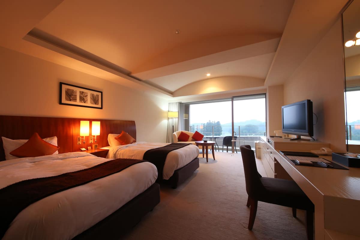 Quarto de hotel com duas camas, uma escrivaninha, uma cadeira, televisão e uma janela grande com vista.