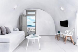 Quarto moderno e minimalista com teto branco curvo, um sofá com almofadas, uma mesa redonda pequena com um vaso, duas cadeiras e uma TV fixada na parede.
