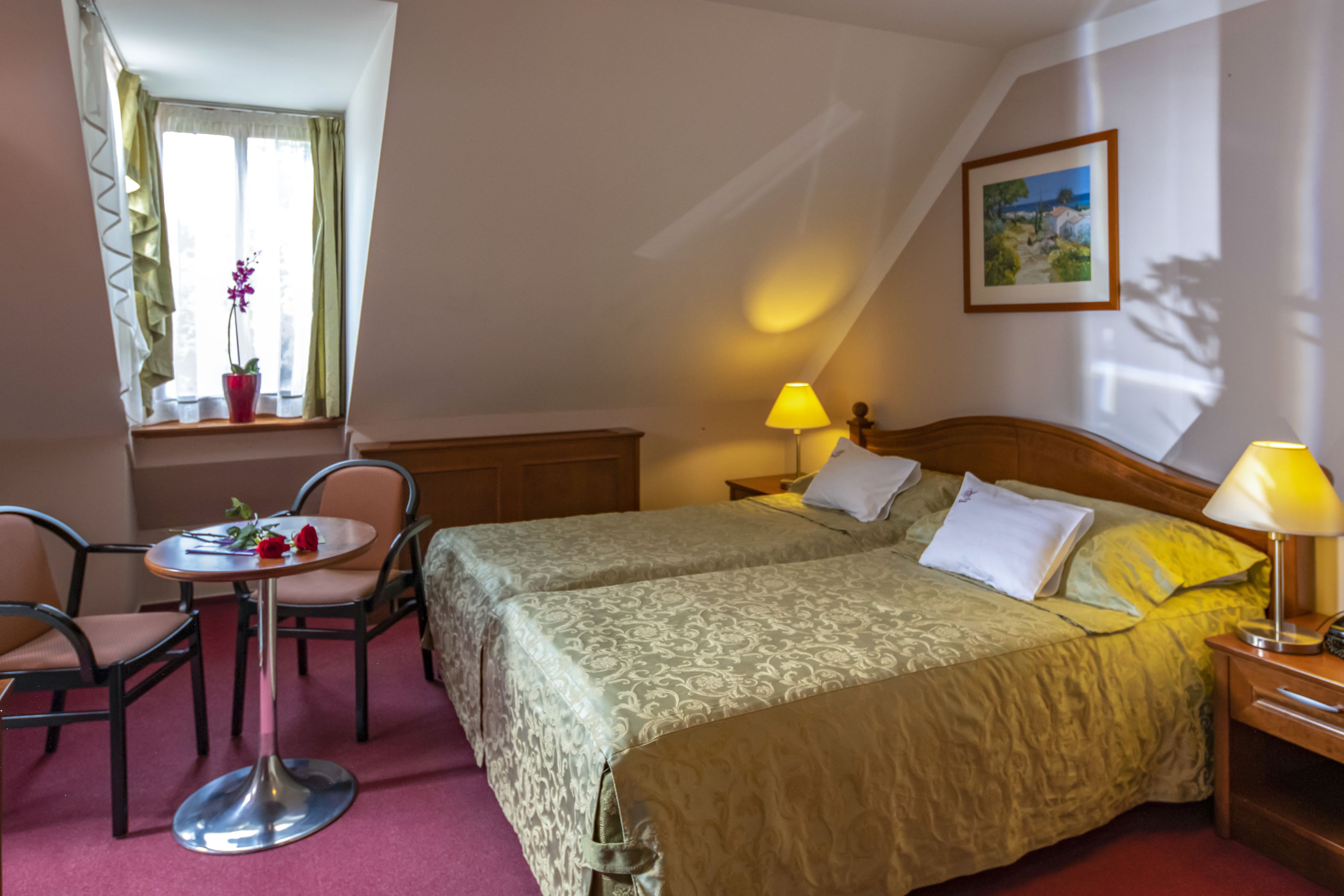 Quarto de hotel com duas camas, uma mesinha com cadeiras, uma janela com cortinas e um quadro na parede.