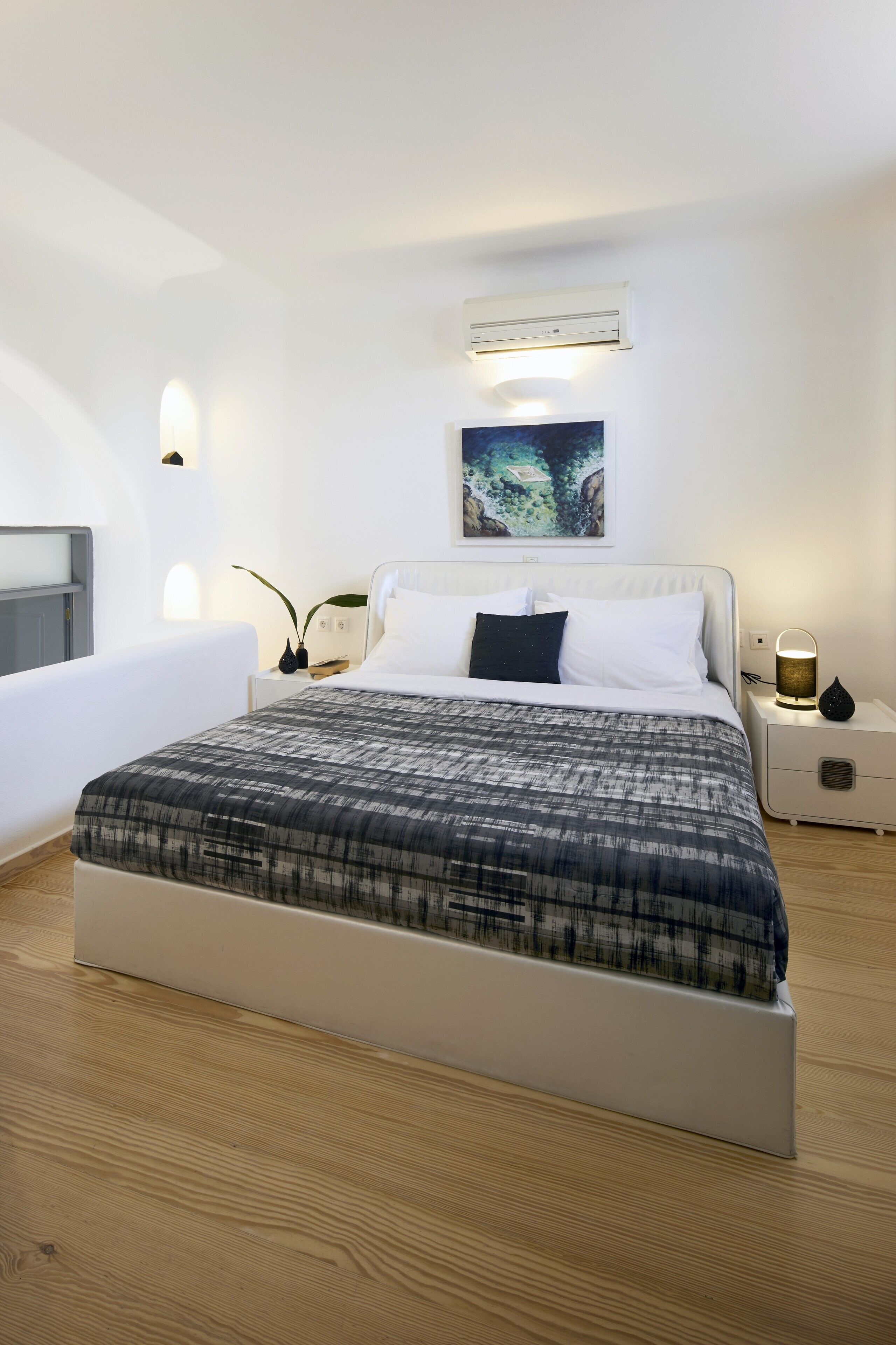 Quarto moderno com uma cama grande, mesa de cabeceira com abajur, ar condicionado na parede e um quadro.