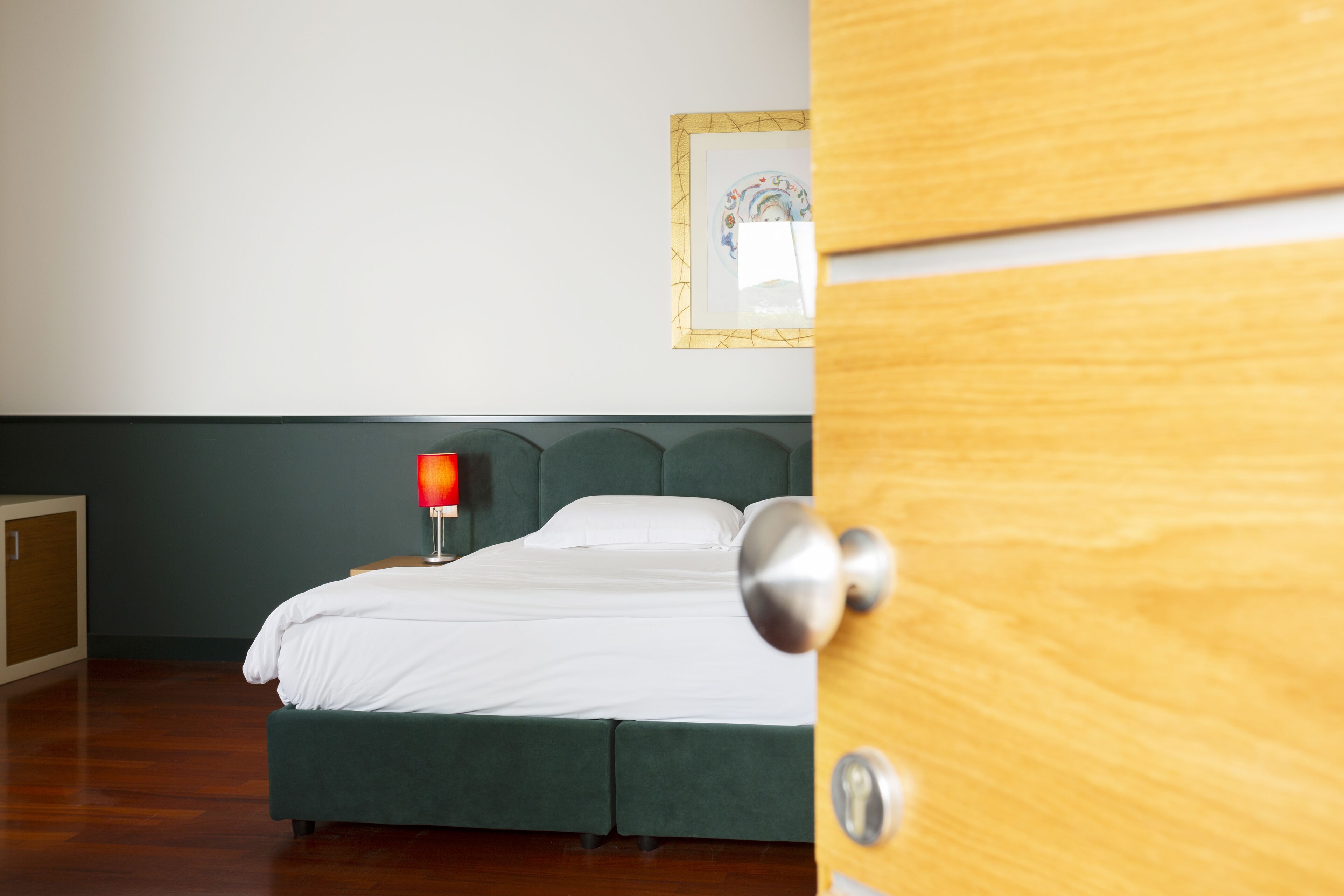 Quarto com cama, mesa de cabeceira com abajur, quadro na parede e porta de madeira.