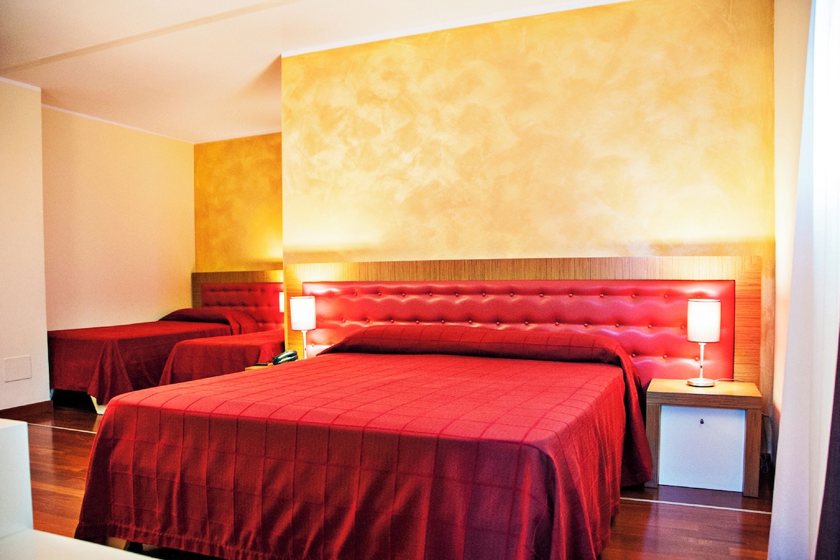 Quarto de hotel com duas camas, cabeceira vermelha e uma pequena mesa de cabeceira com abajur.