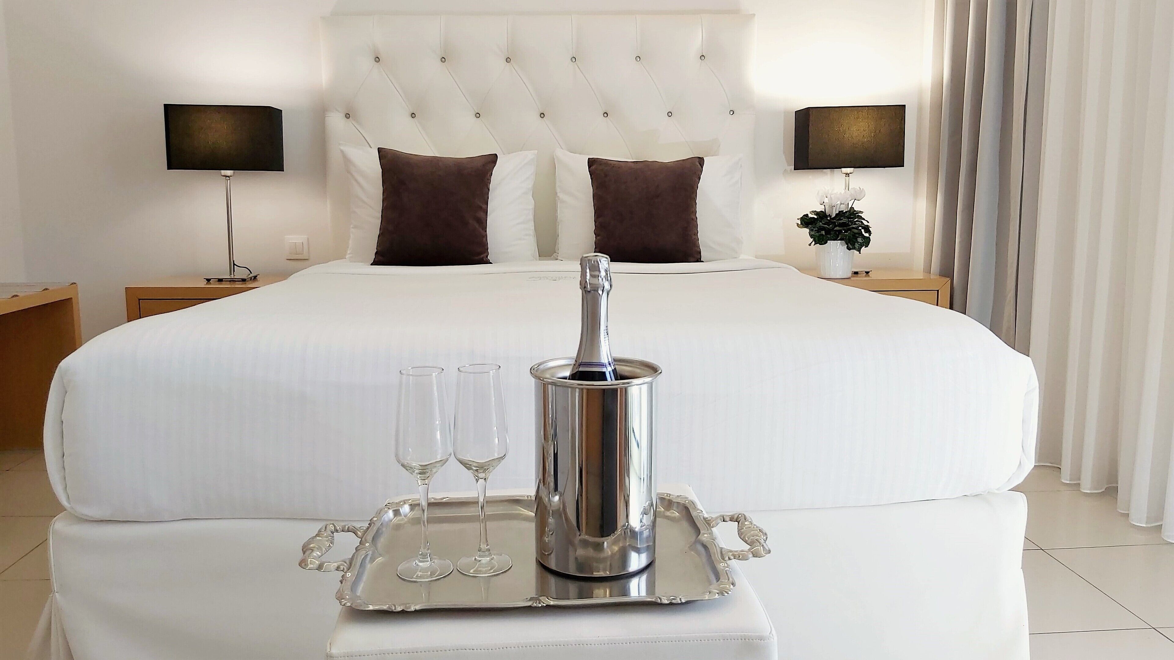 Cama bem arrumada com uma garrafa de champanhe e duas taças em um tabuleiro, cabeceira estofada e duas luminárias de mesa.