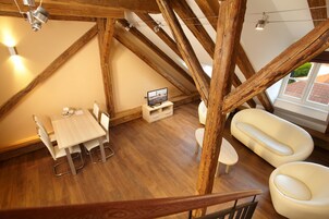 Um quarto estilo loft com vigas de madeira aparentes, uma sala de jantar com mesa e cadeiras, um sofá branco e uma televisão sobre um móvel baixo.