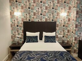 Uma cama bem arrumada com almofadas brancas e azuis, uma cabeceira escura e um papel de parede estampado atrás dela.
