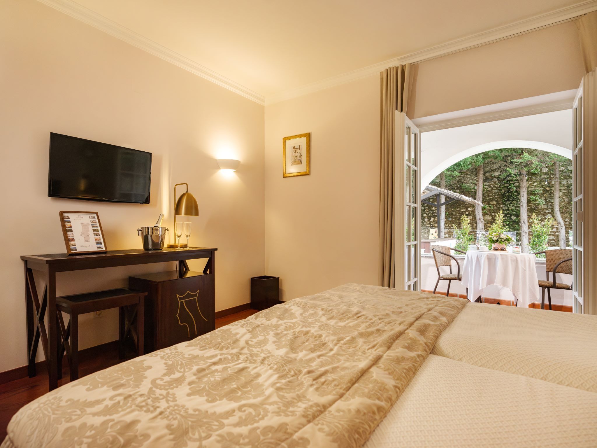 Quarto de hotel com cama, escrivaninha com abajur, televisão e vista para um pátio externo com mesa e cadeiras.