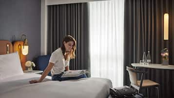 Quarto de hotel com cama, mesa de cabeceira, abajur, cadeira, mesa e janela com cortinas.