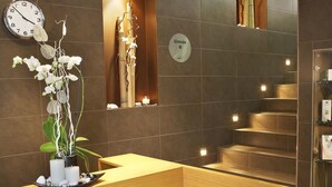 Uma área de recepção moderna com um balcão de madeira, um banco com almofadas florais, um relógio e um arranjo decorativo de orquídeas.