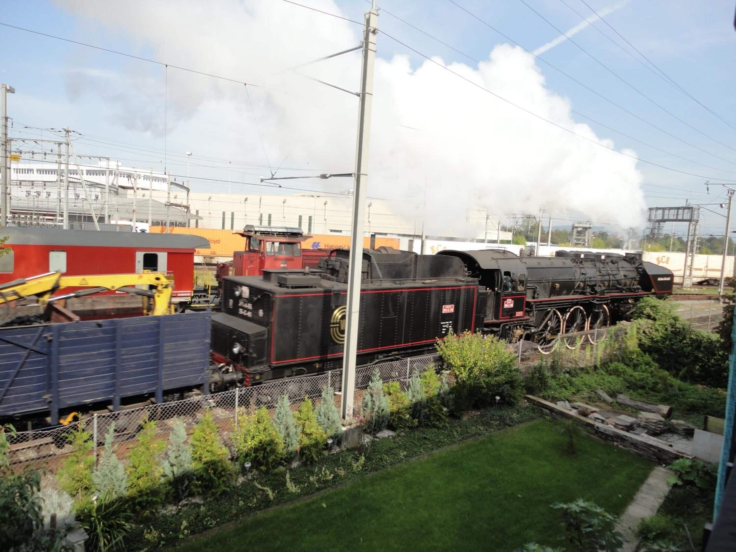 Uma estação de trem com várias locomotivas, um edifício vermelho e estruturas industriais ao fundo.
