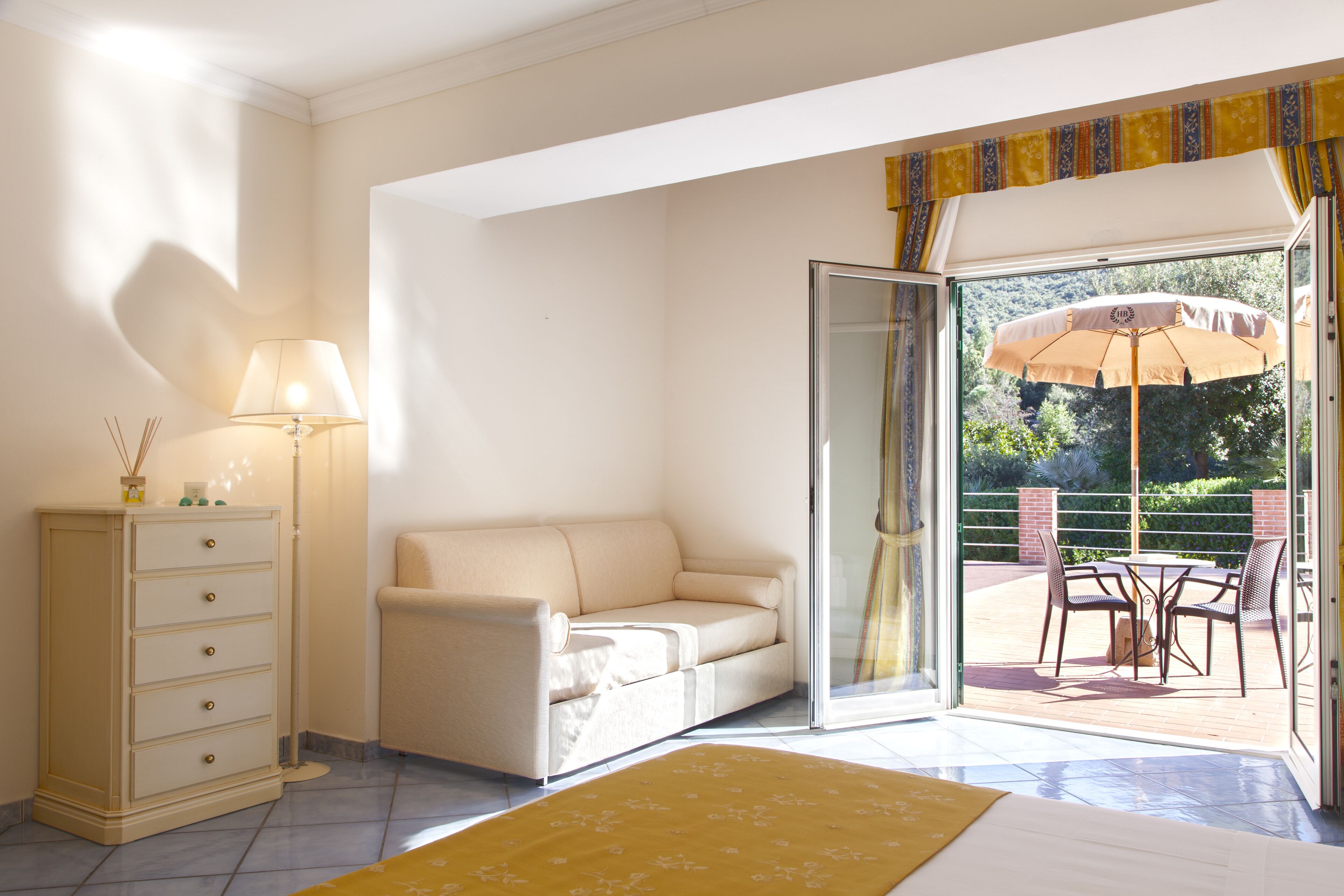 Quarto com cama, sofá, luminária, cômoda e porta de vidro deslizante que dá para um pátio externo com cadeiras e guarda-sol.