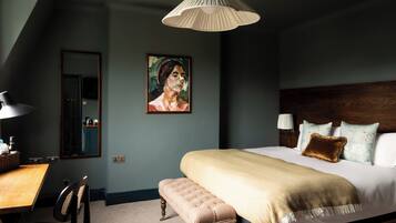 Quarto com uma cama grande, cabeceira de madeira, mesa de cabeceira, luminária, um banquinho e um quadro na parede.