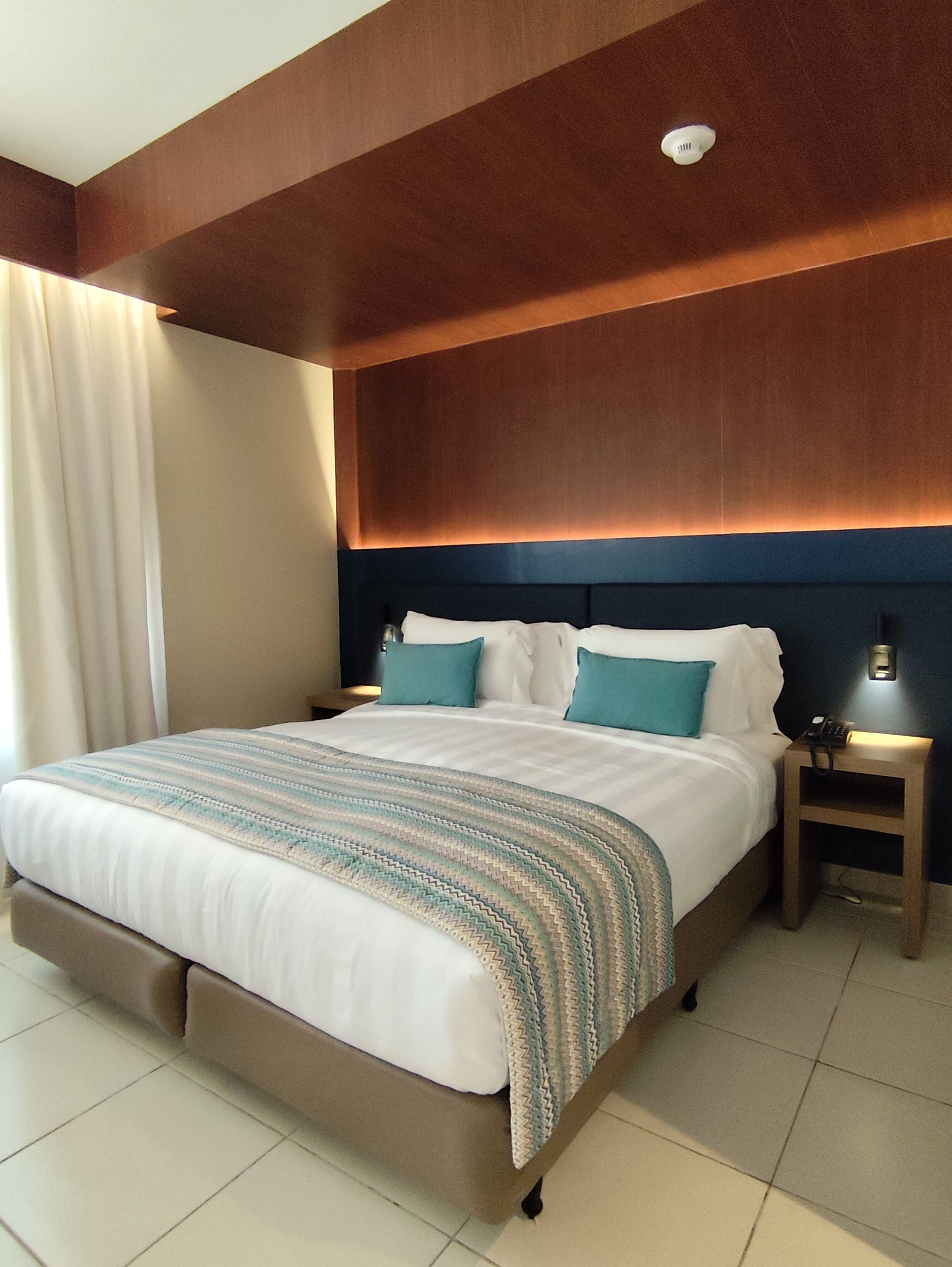 Quarto de hotel moderno com uma cama grande, uma mesa de cabeceira com telefone e uma luminária fixada na parede.