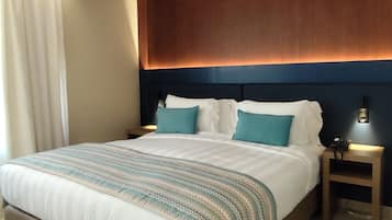 Quarto de hotel moderno com uma cama grande, uma mesa de cabeceira com telefone e uma luminária fixada na parede.