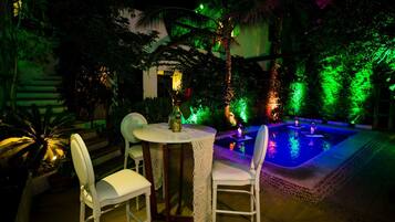 Uma área de piscina com mesa e cadeiras, cercada por vegetação exuberante e iluminação decorativa.