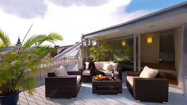 Terraço no telhado com mobiliário de vime, uma mesa de jantar com frutas e vinho, e vista para uma torre de igreja ao fundo.
