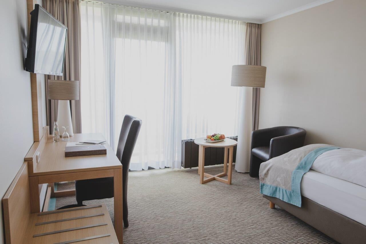 Quarto de hotel com uma cama, uma escrivaninha com cadeira, uma mesinha de cabeceira com abajur e uma televisão na parede.