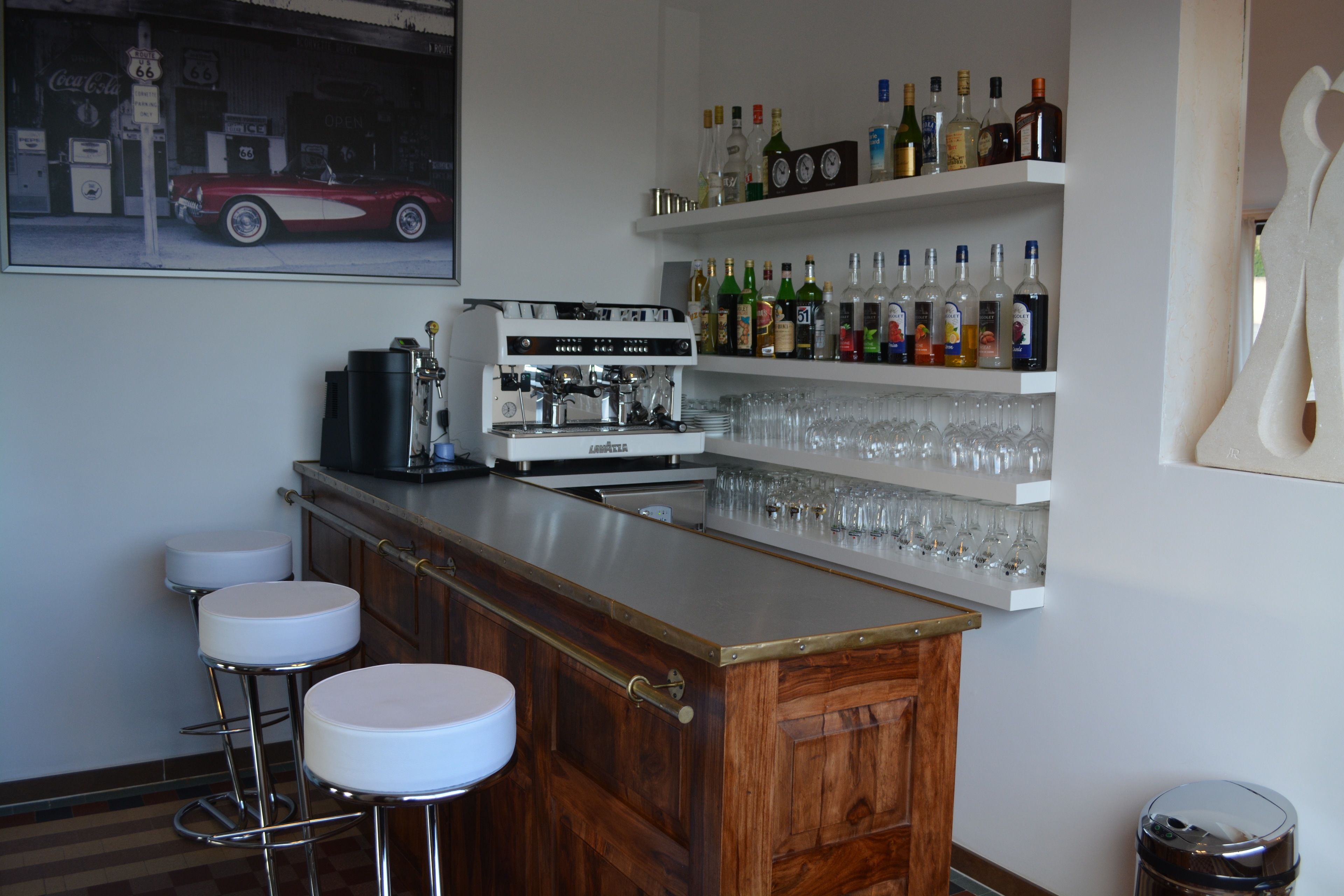 Uma área de bar com uma máquina de café, banquetas, prateleiras com garrafas de bebidas e um relógio fixado na parede.