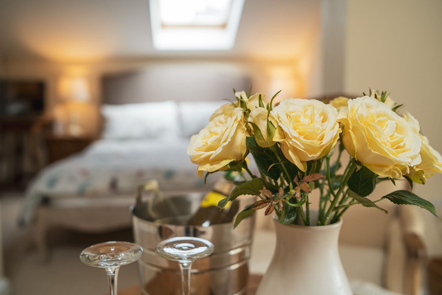 Quarto com cama, mesa de cabeceira com um vaso de rosas amarelas e duas taças de vinho.