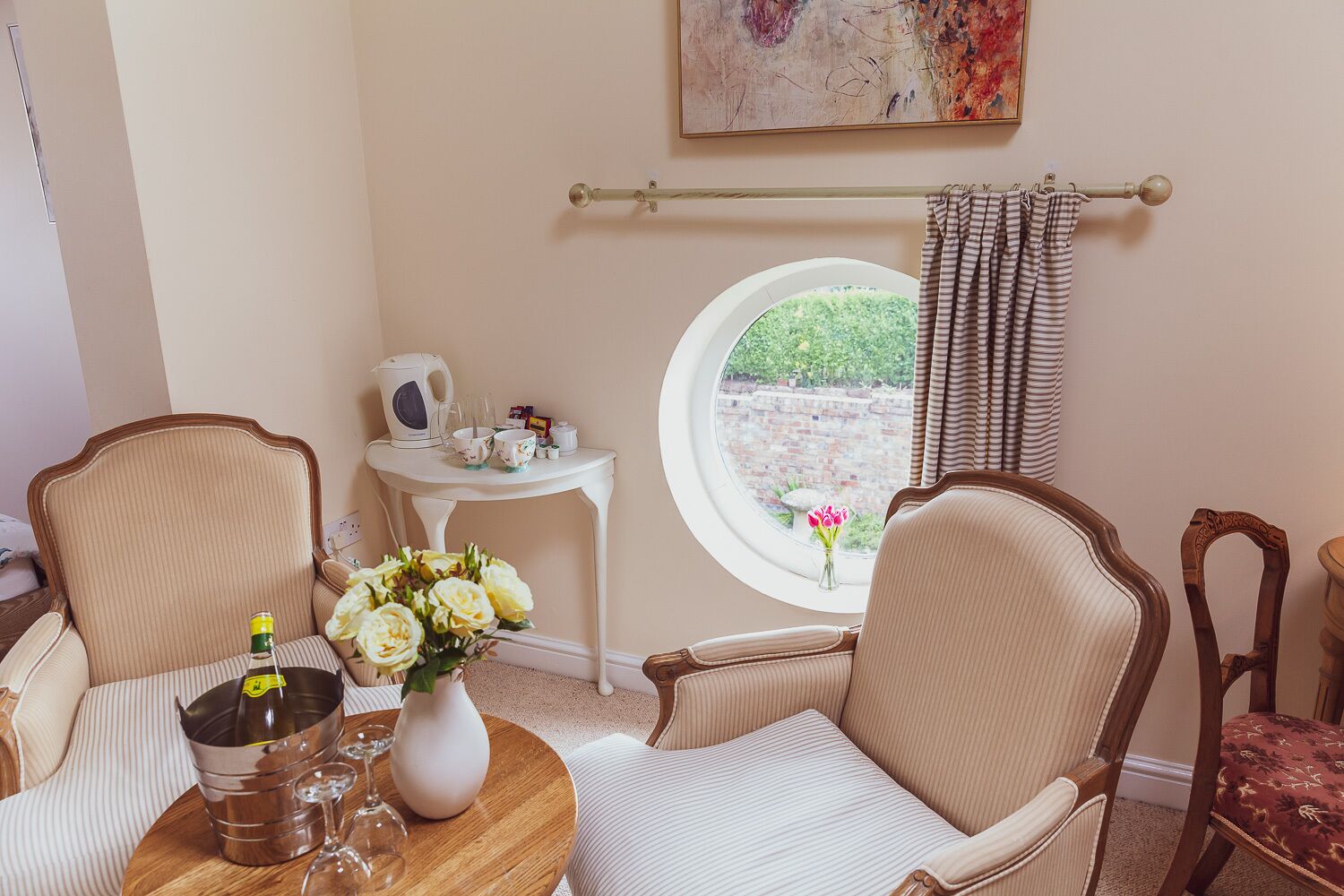 Quarto aconchegante com duas poltronas estofadas, uma mesinha redonda, uma garrafa de champanhe, taças e um vaso de flores. Há uma janela com vista para o verde e um muro de tijolos lá fora.