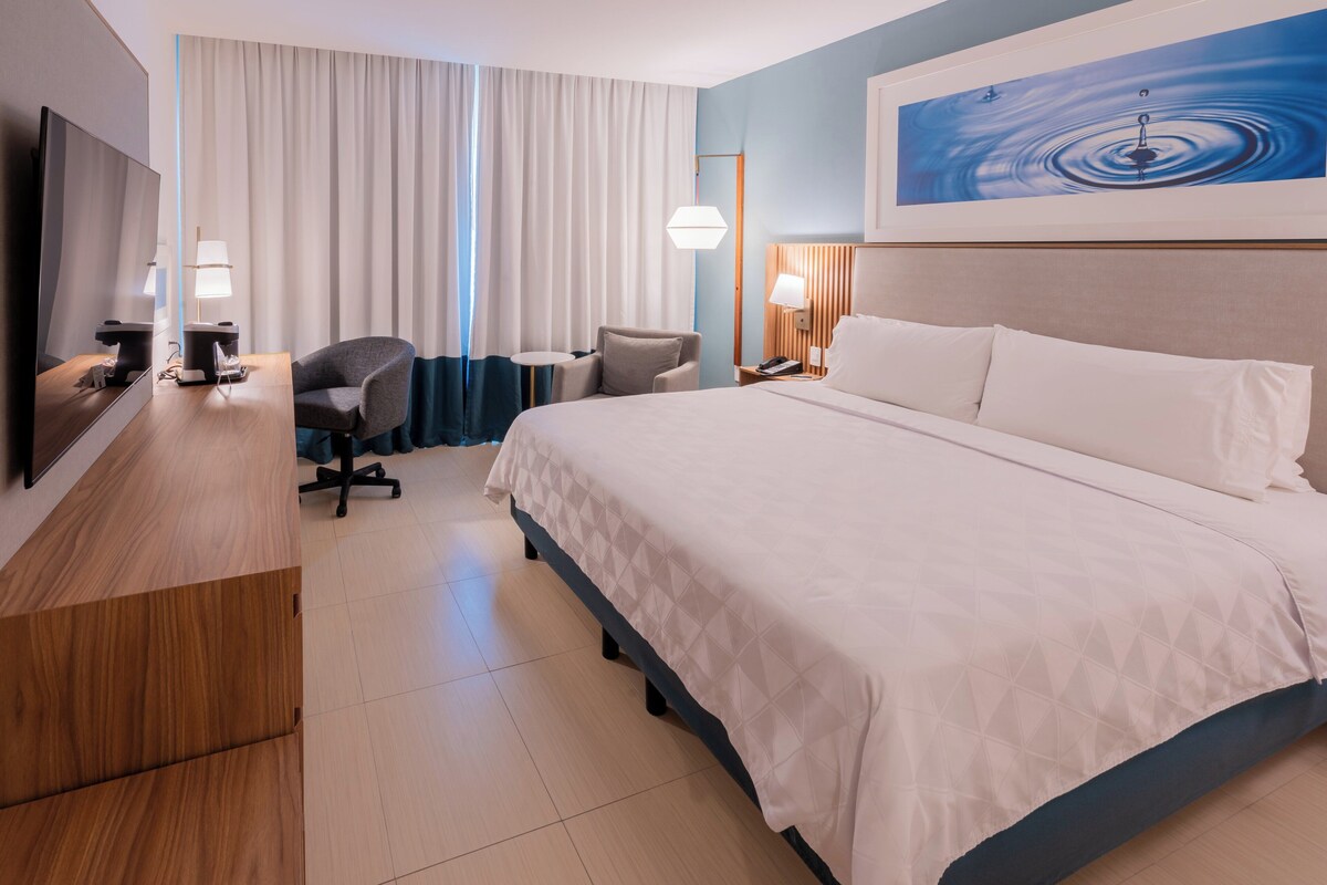 Quarto de hotel com uma cama grande, uma escrivaninha com televisão de tela plana, uma cadeira e uma mesa pequena.
