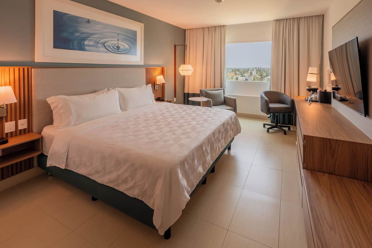Quarto de hotel com uma cama grande, uma escrivaninha com cadeira, televisão e janela com cortinas.