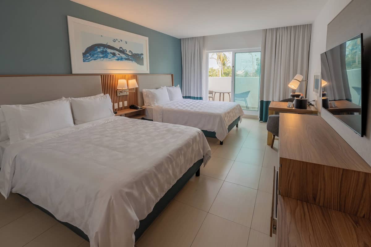 Quarto de hotel com duas camas, uma escrivaninha com cadeira, televisão e varanda com vista para palmeiras.