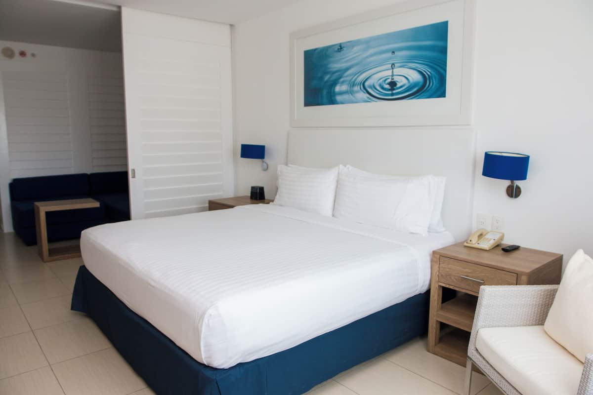 Quarto de hotel com uma cama grande, mesa de cabeceira com abajur, uma cadeira e um quadro na parede.