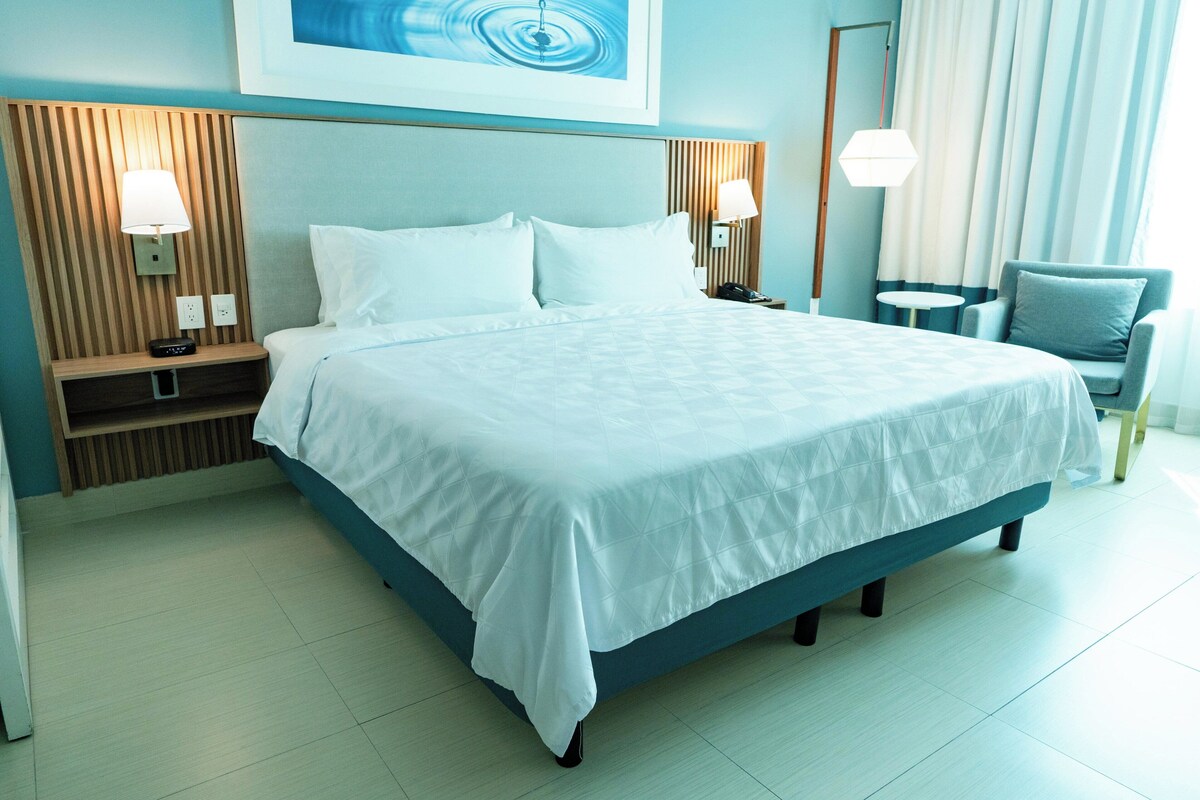 Quarto de hotel com uma cama grande, abajures, uma cadeira e vista para o exterior.