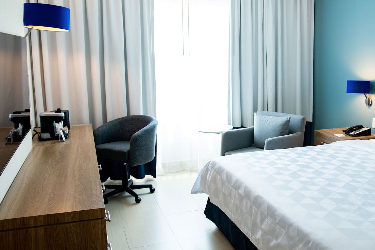 Quarto de hotel com cama, escrivaninha com luminária, cadeira e mesa de cabeceira com telefone.