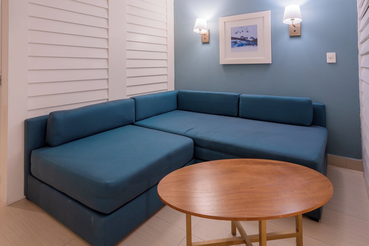 Um sofá azul em formato de L com uma mesa de madeira na frente, persianas brancas à esquerda e luminárias fixadas na parede.