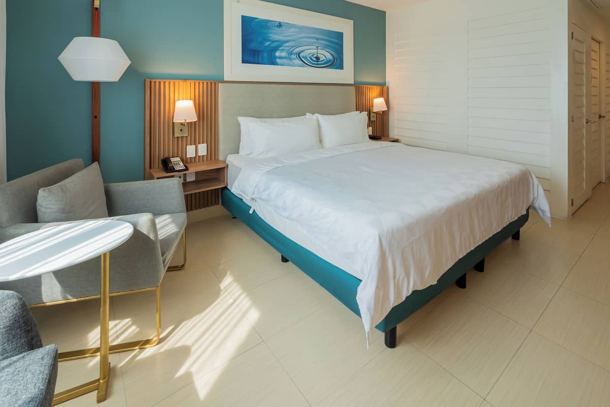 Quarto de hotel com cama, escrivaninha, cadeira, abajur e um quadro na parede.