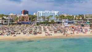 Uma praia com vários guarda-sóis e pessoas. Ao fundo, o hotel 'Hotel Riu San Francisco'.