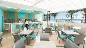 Uma área de bar ao ar livre com assentos, um bartender e vista para a praia.