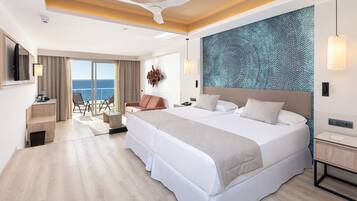 Quarto de hotel moderno com uma cama grande, um ventilador de teto, vista para o oceano e um espaço de estar com uma cadeira e mesa.
