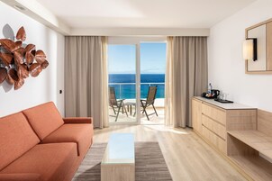 Quarto de hotel moderno com sofá, área de refeições com mesa e cadeiras, cozinha e varanda com vista para o oceano.