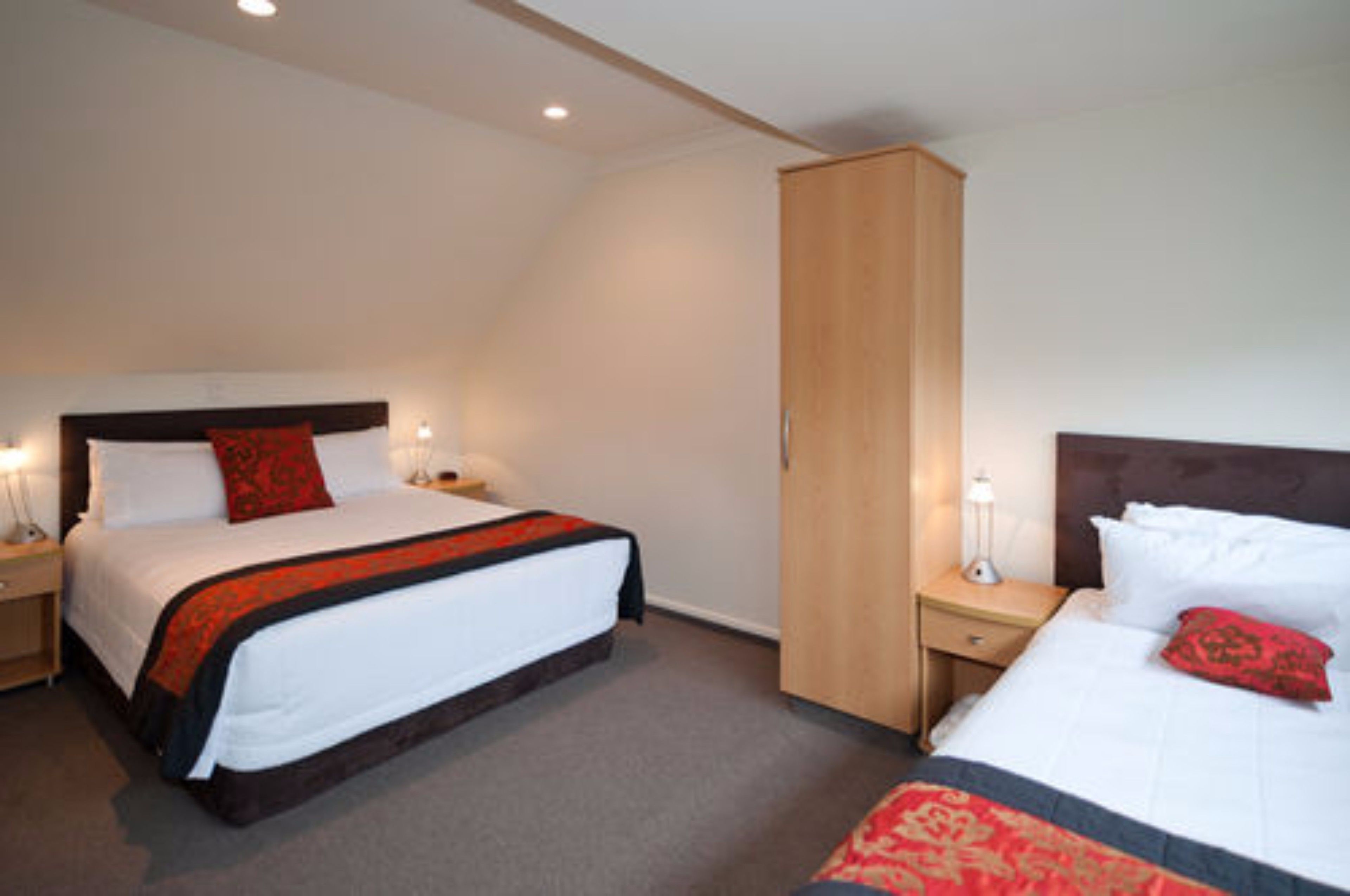 Quarto de hotel com duas camas, guarda-roupa, mesas de cabeceira com luminárias e chão coberto por carpete.