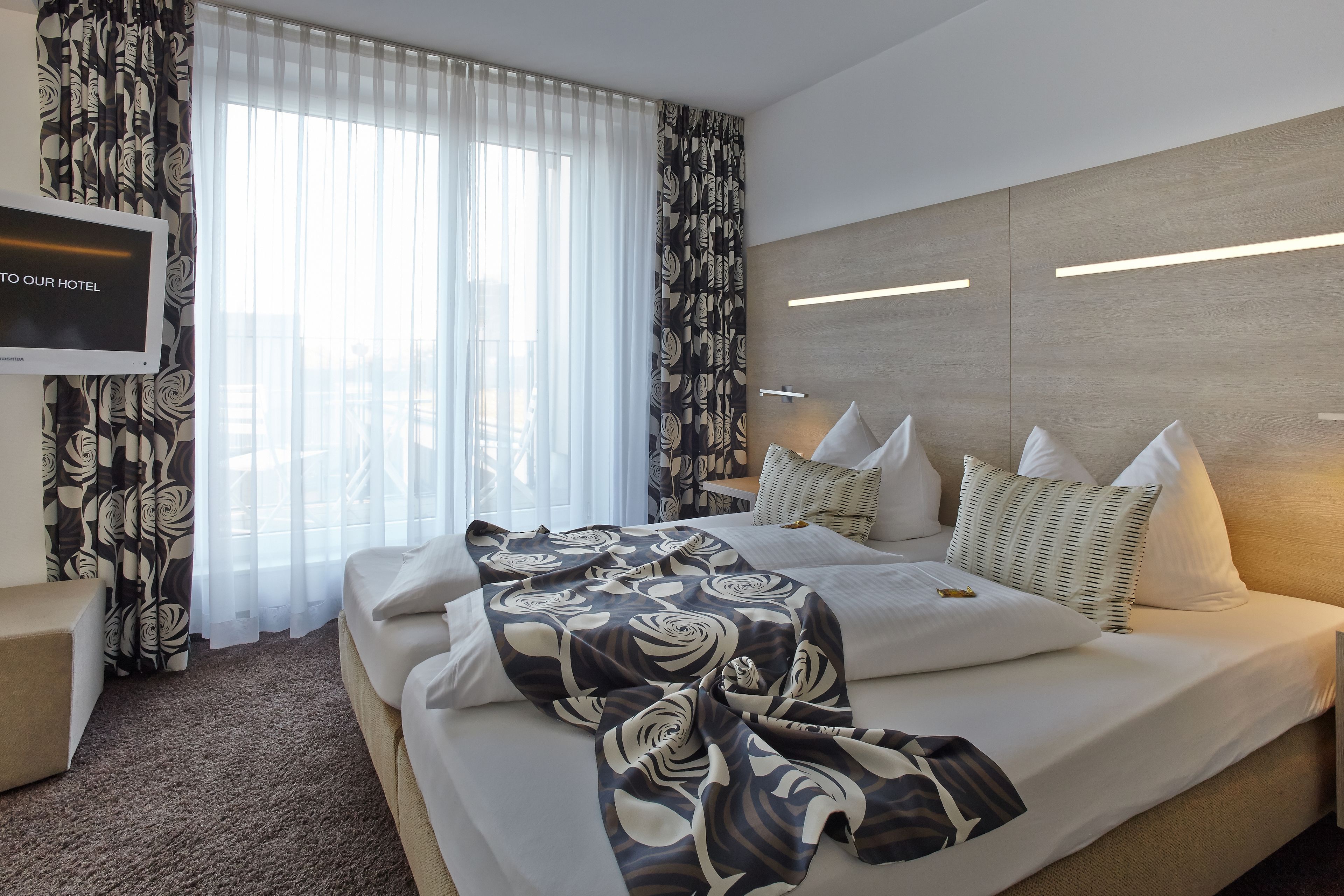 Quarto de hotel com uma cama grande, televisão de tela plana, escrivaninha e vista da cidade por cortinas finas.