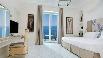 Quarto de hotel com cama, escrivaninha com computador, cadeira, televisão e varanda com vista para o mar.