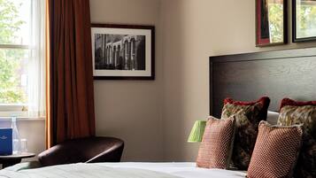 Quarto de hotel com cama, uma cadeira, janela com cortinas, quadro na parede e abajur sobre uma mesinha de cabeceira.