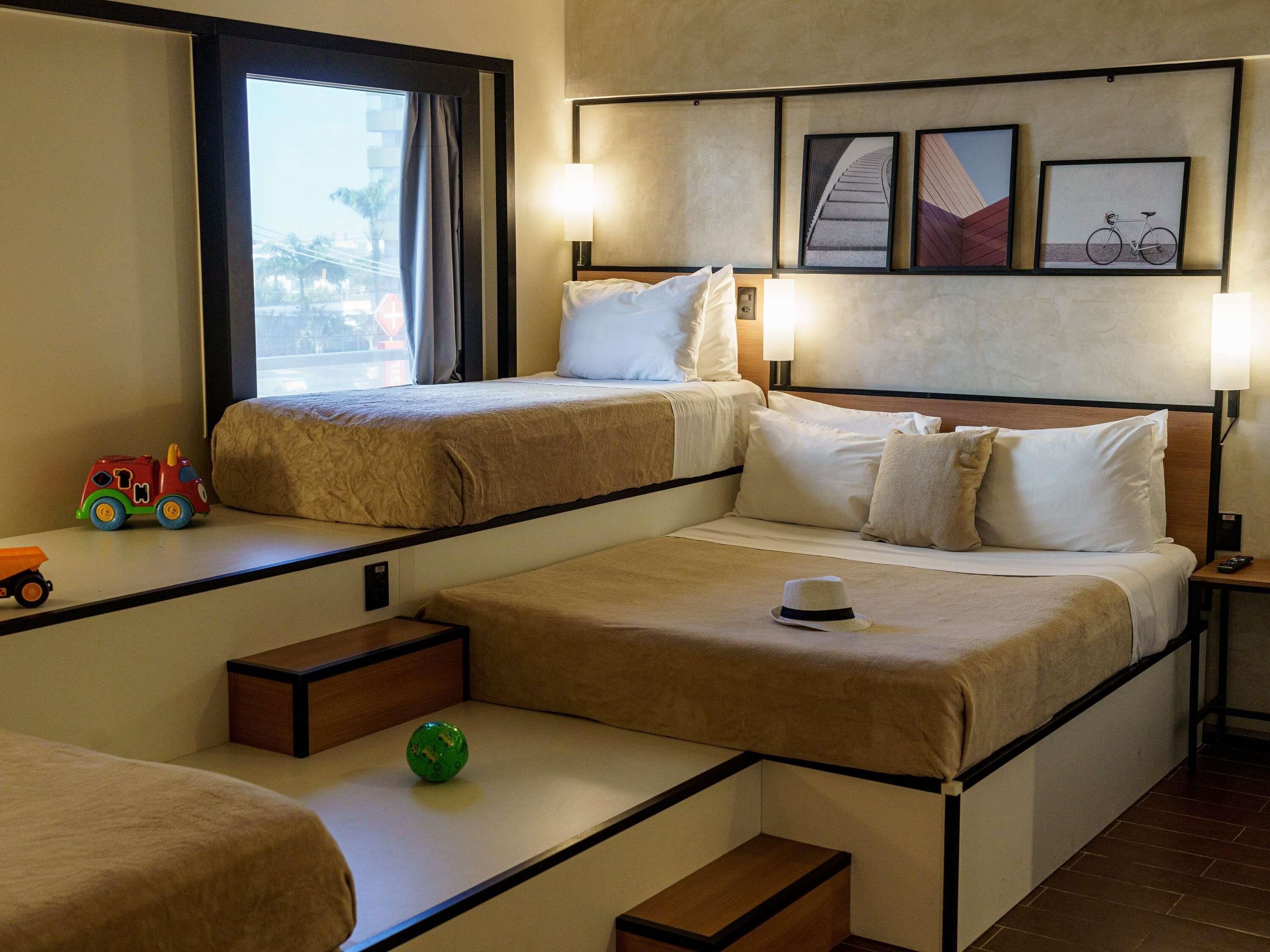 Quarto de hotel com duas camas, janela com cortinas, abajur, quadros na parede e um carrinho de brinquedo no parapeito da janela.