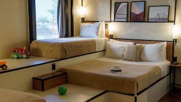 Quarto de hotel com duas camas, janela com cortinas, abajur, quadros na parede e um carrinho de brinquedo no parapeito da janela.