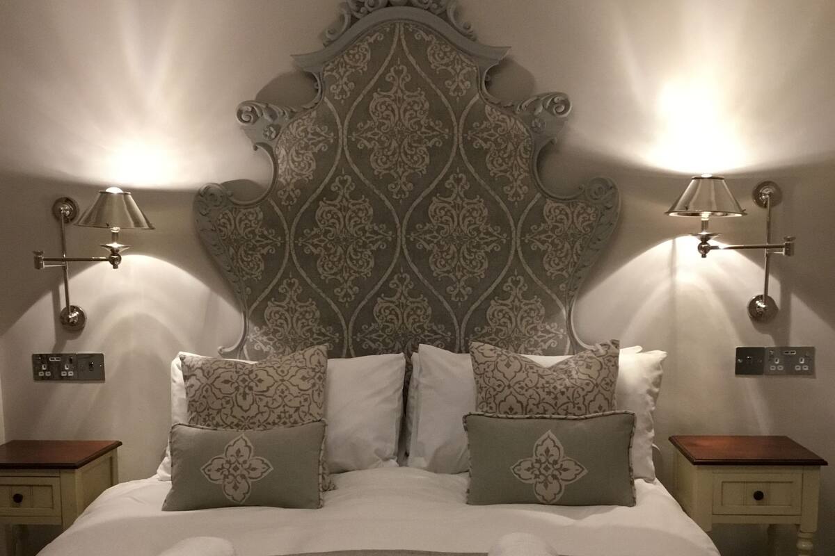 Cama bem arrumada com cabeceira decorativa, duas mesas de cabeceira com luminárias e painel de controle fixado na parede.