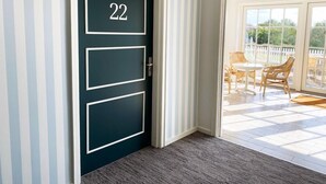 Uma porta azul escura com o número 22, dando acesso a uma varanda com cadeiras de vime e um cercado branco.