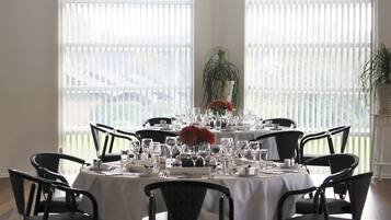 Uma sala de jantar com uma mesa redonda posta para seis pessoas, rodeada por cadeiras pretas, sob iluminação pendente, com grandes janelas e vista para o exterior.